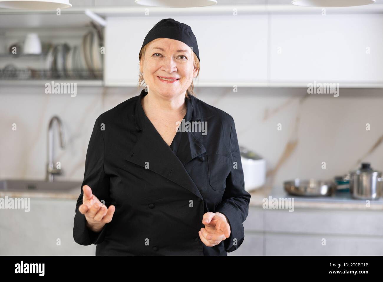 Happy mature female cook in uniform standing and showing her kitchen ...