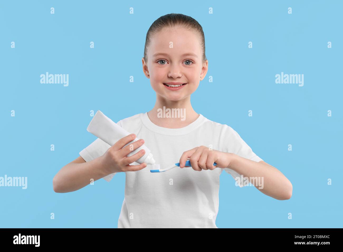 Happy girl squeezing toothpaste from tube onto toothbrush on light blue ...