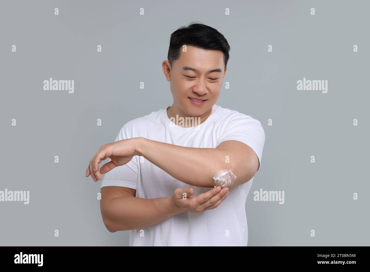 Handsome man applying body cream onto his elbow on light grey ...