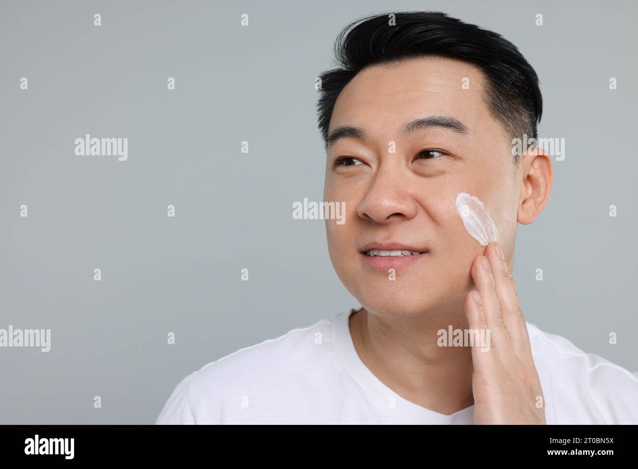 Handsome man applying cream onto his face on light grey background ...
