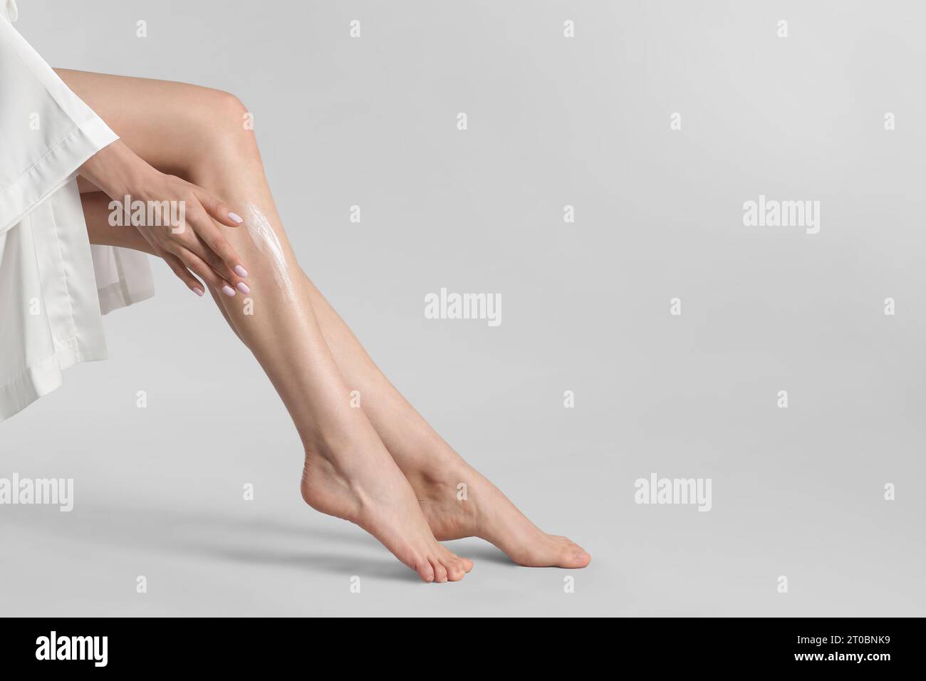 Woman applying body cream onto her smooth legs on light grey background ...