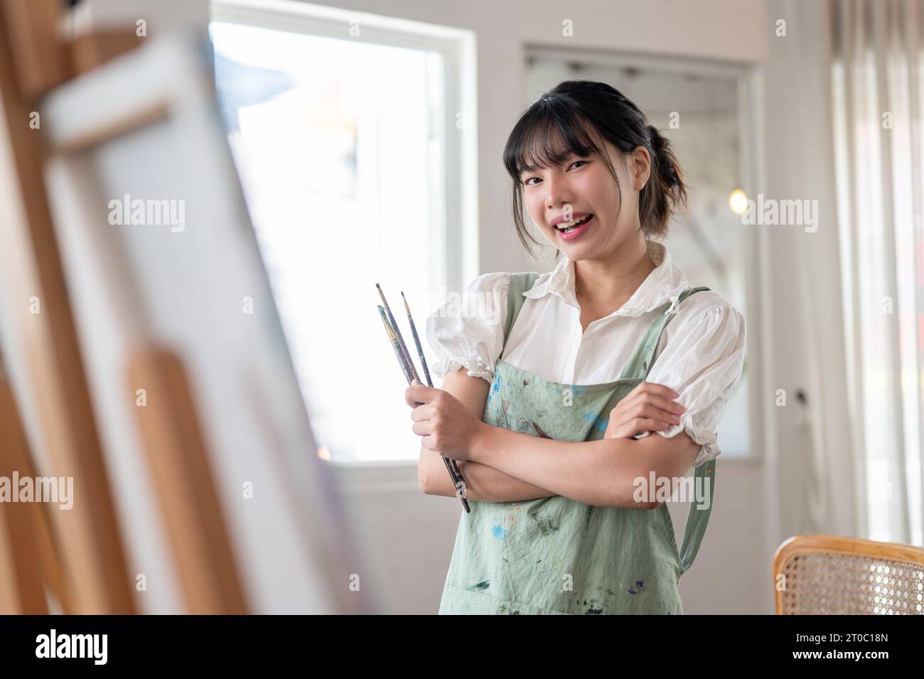 A cheerful and creative young Asian female artist in an apron smiles at ...