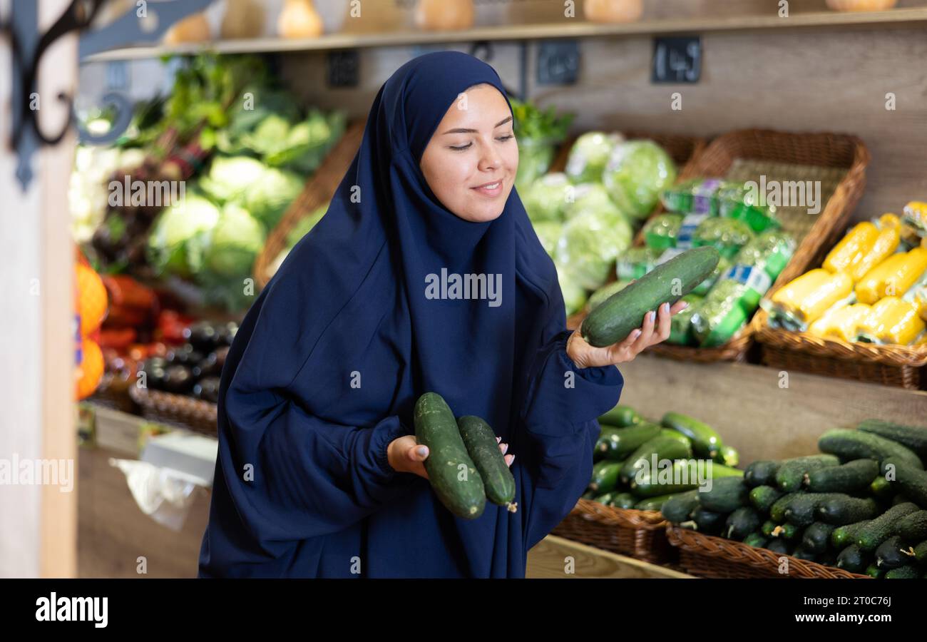 Young muslim woman in abaya and chador choosing cucumbers in store ...