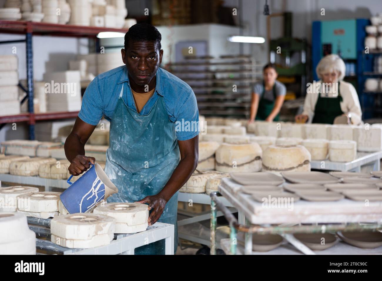 African american worker pours slip clay mass from jug into plaster mold ...