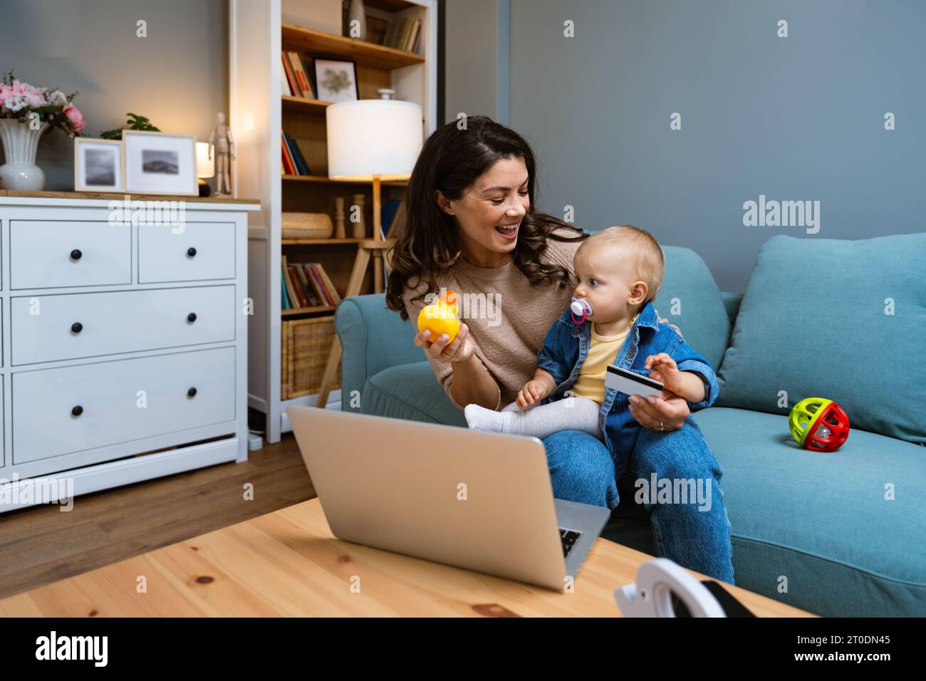 Young happy mother holding her baby while using credit card and laptop for online banking at ...