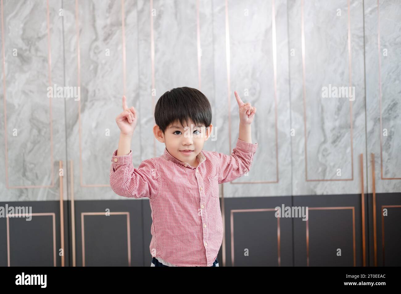 An Asian boy stands and smiles, raising both hands and pointing upwards ...