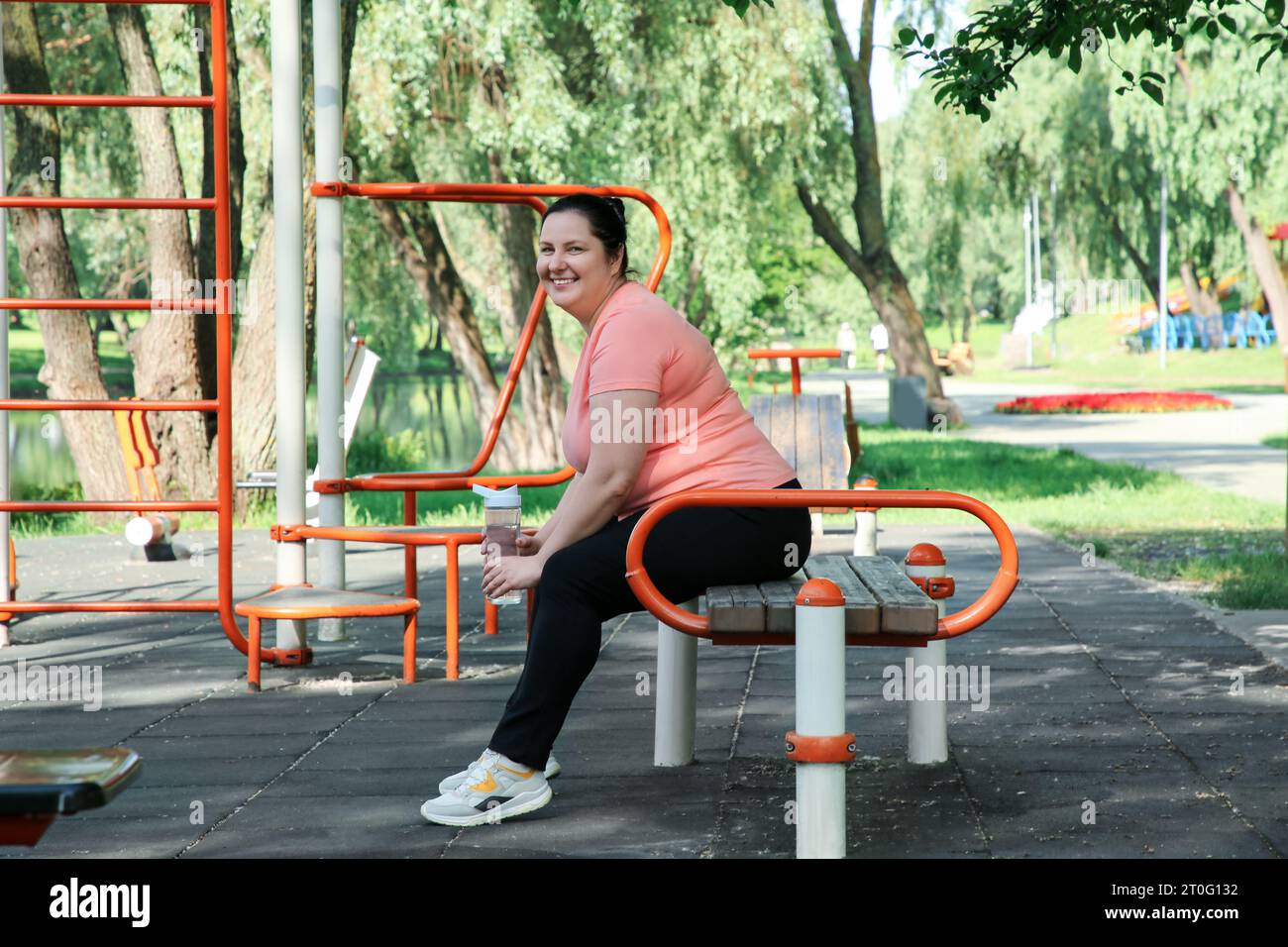 Overweight woman with bottle of water sitting on bench of sports ground ...