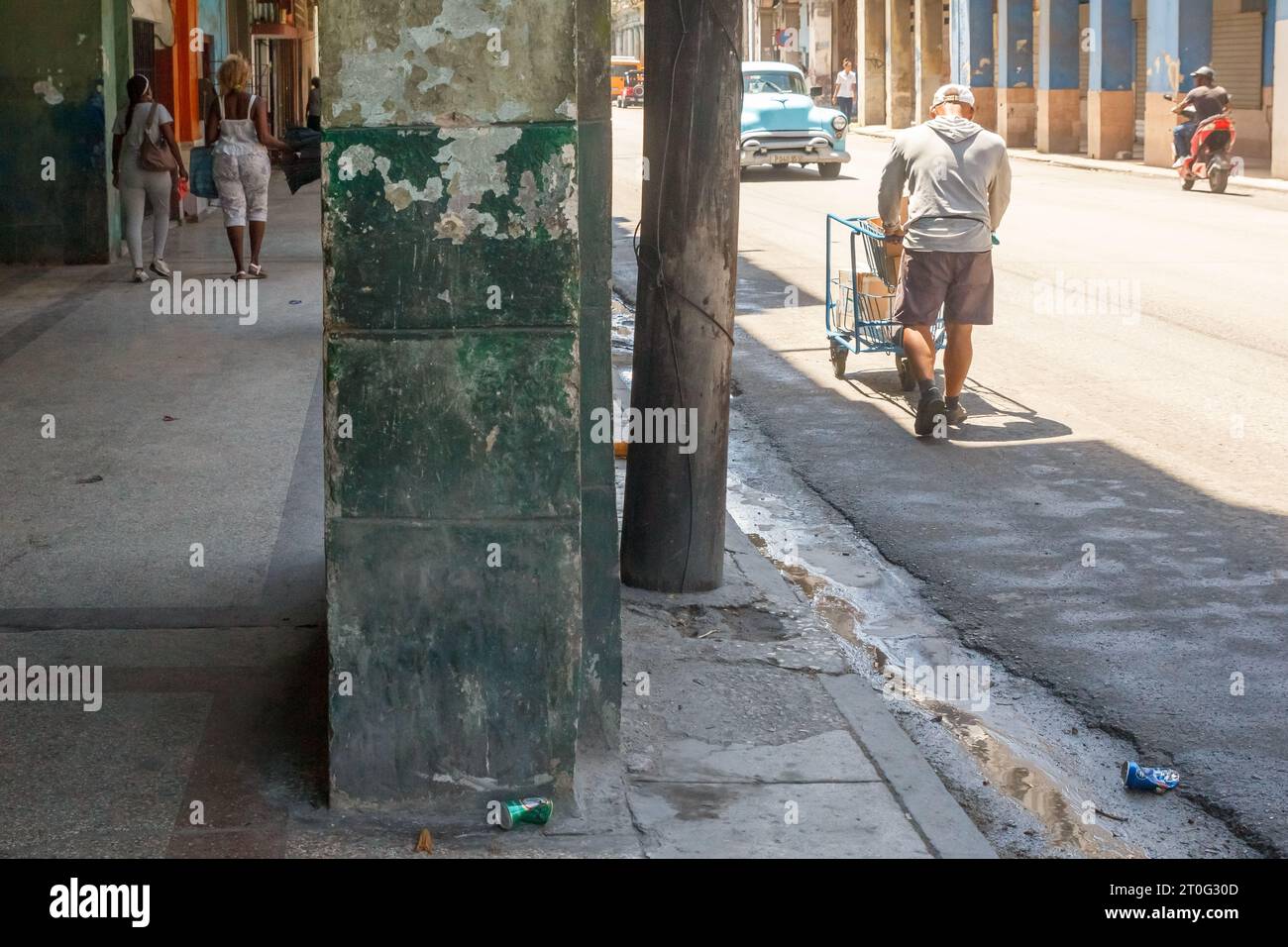 A Cuban man pushes a small cart on a city street. Stagnant dirty water ...
