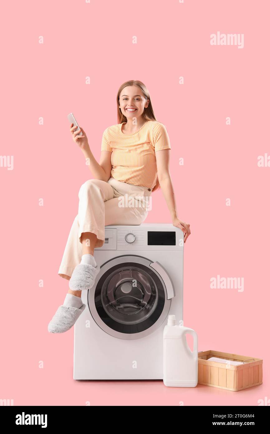Pretty young woman with mobile phone sitting on washing machine against ...