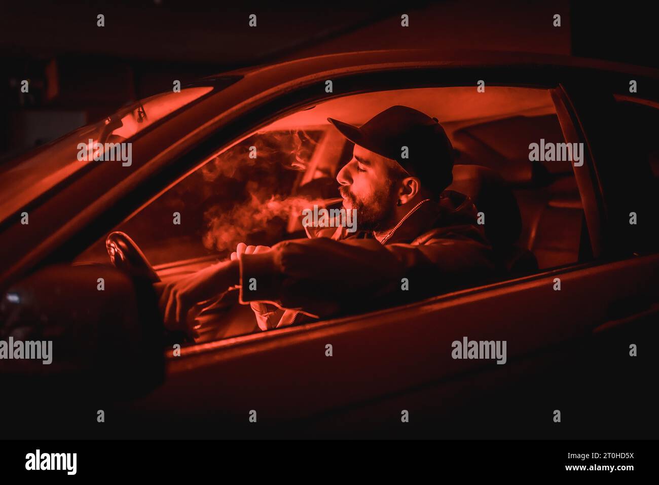 Man in a cap driving car smoking at night in a garage lit with a red ...