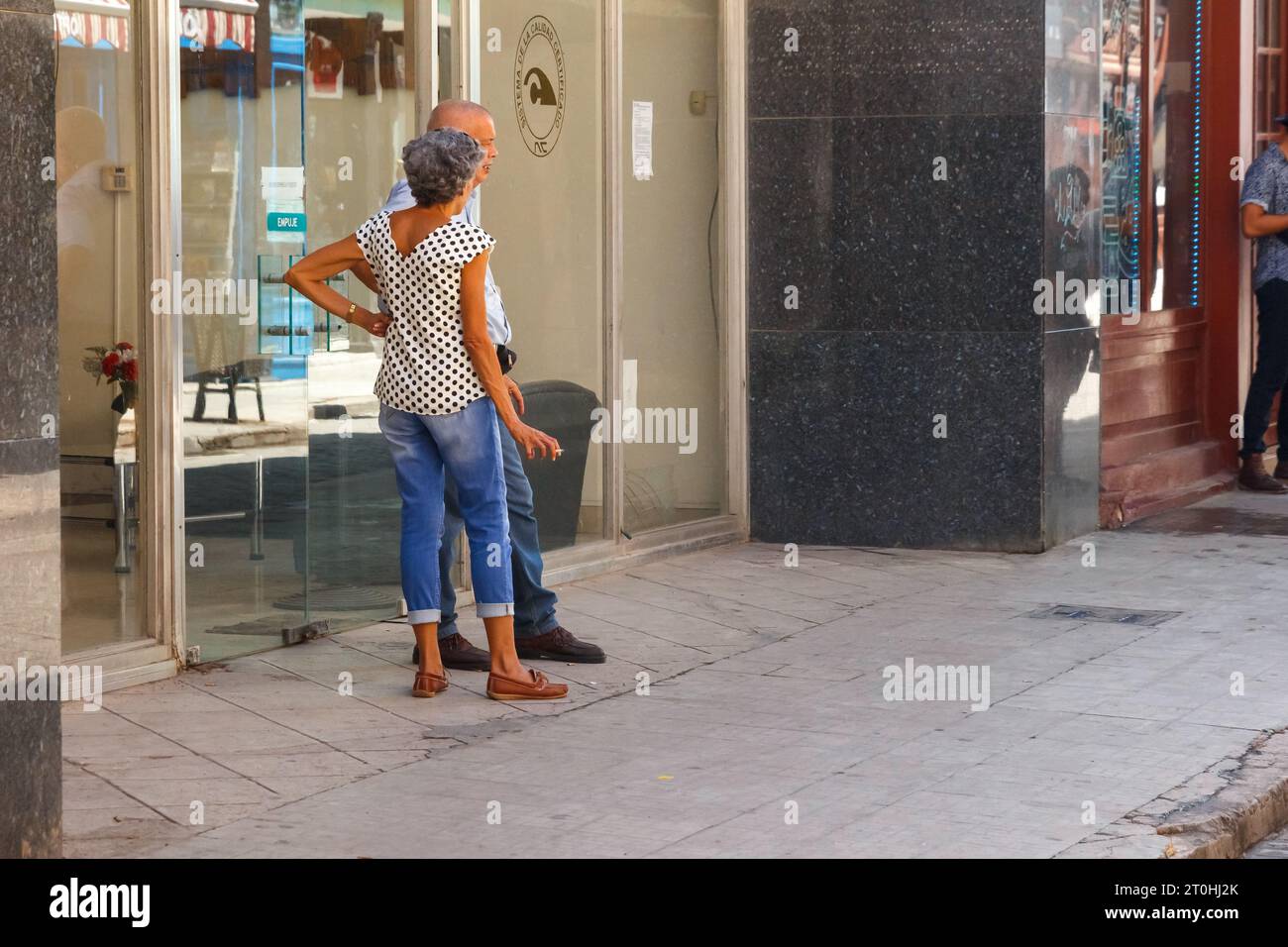 Two Cuban people stand and talk outside a business. The glass of the ...