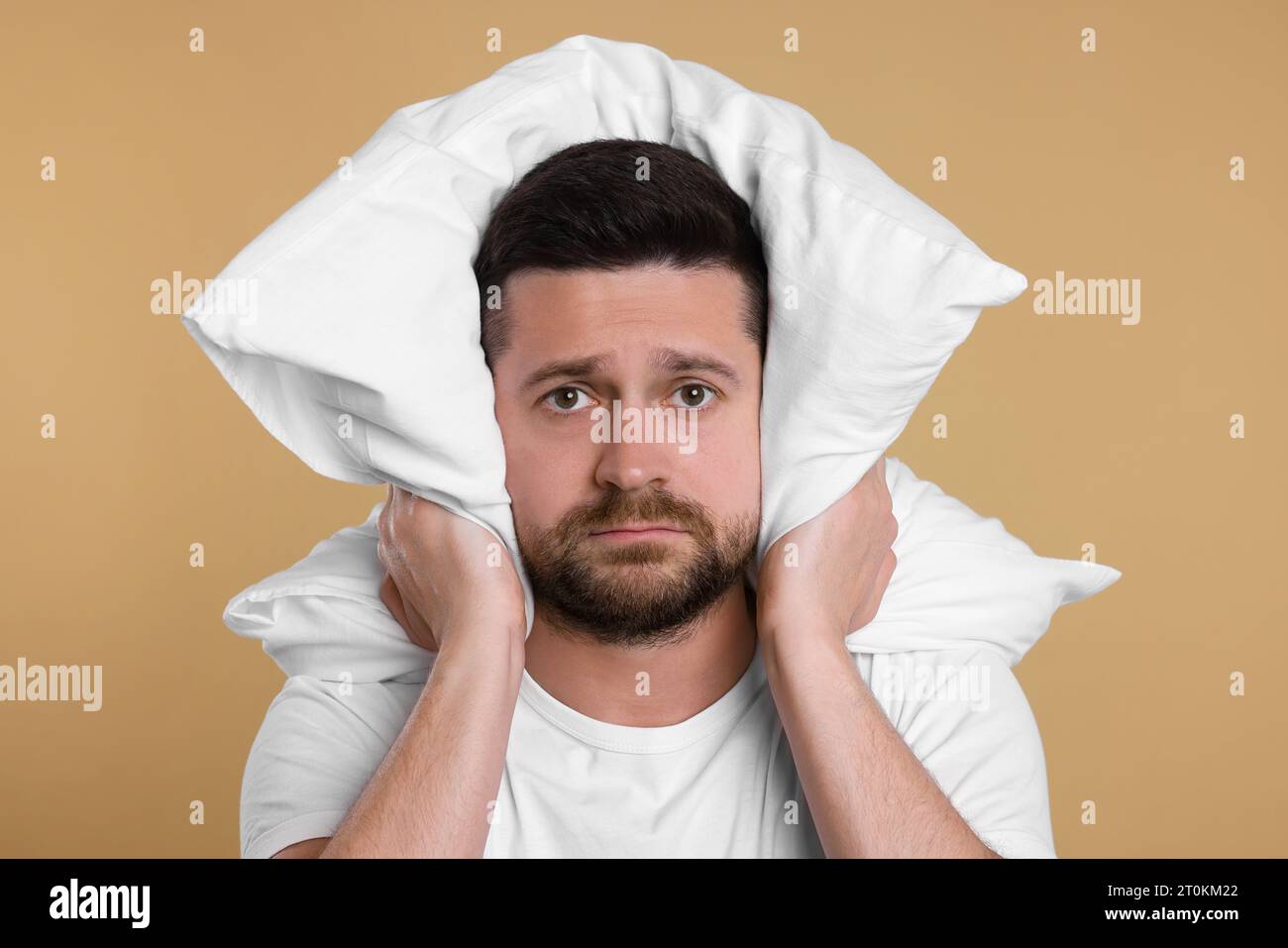 Unhappy man covering ears with pillow on beige background. Insomnia ...