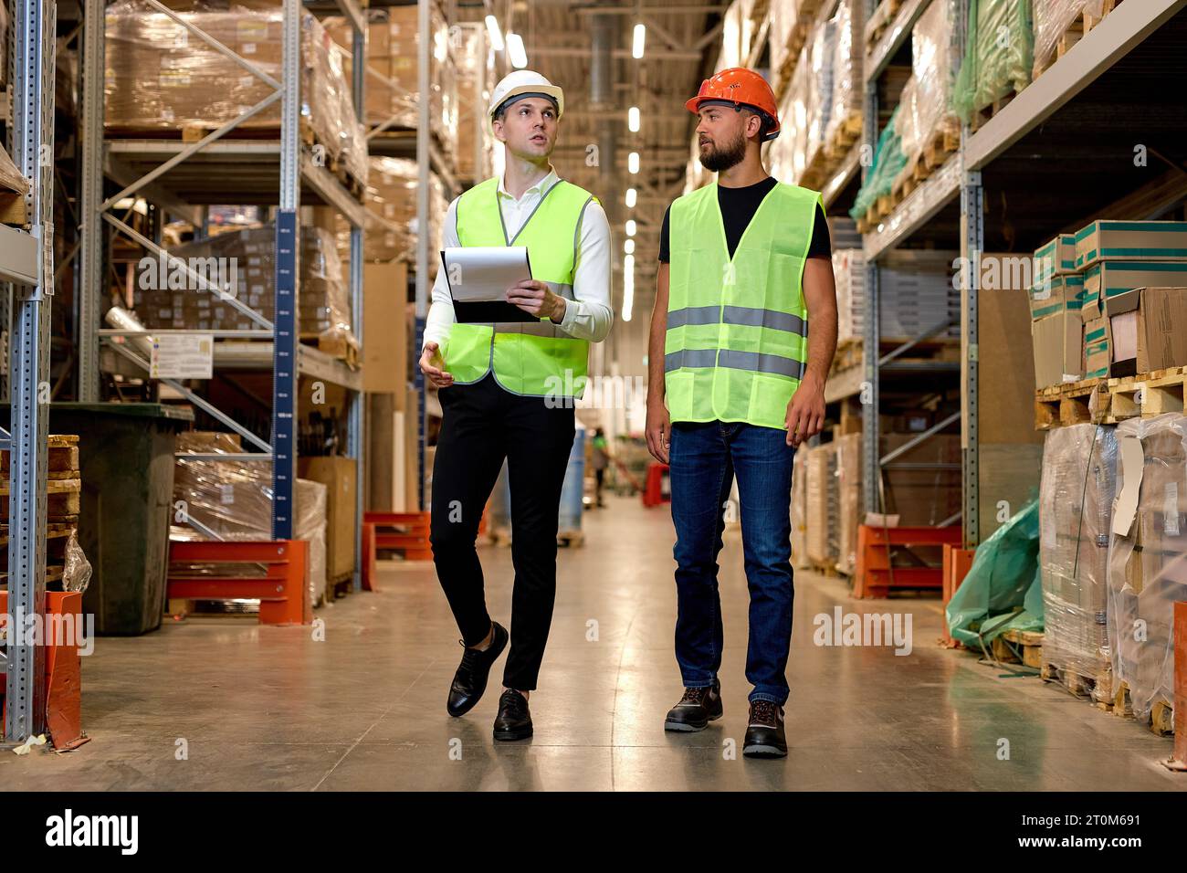Caucasian engineers in helmets and uniform, team checking goods and supplies on shelves in ...