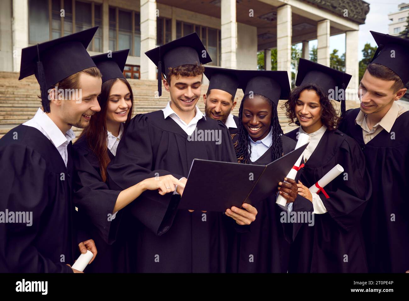 Happy graduate students in black graduation robes looking to diploma of ...