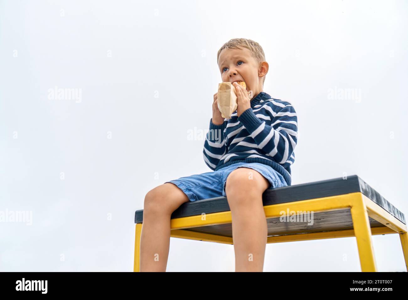 Hungry little boy with hood eats sandwich sitting on table on misty day ...