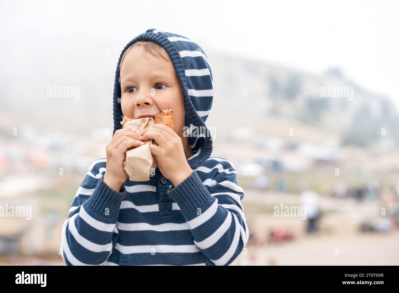 Hungry little boy with hood eats sandwich on misty day. Cute child ...