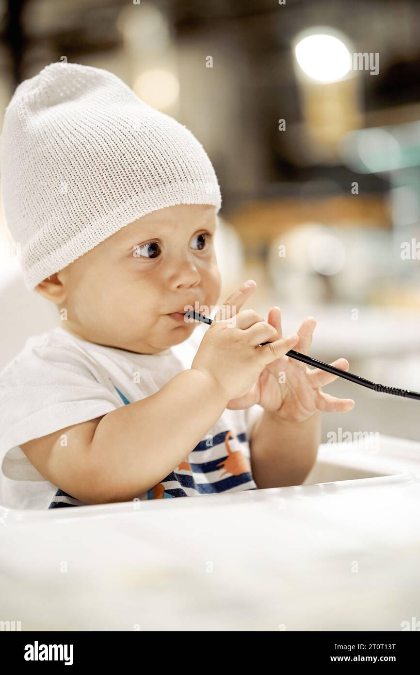 Funny baby holds straw in mouth sitting in high chair closeup. Little ...