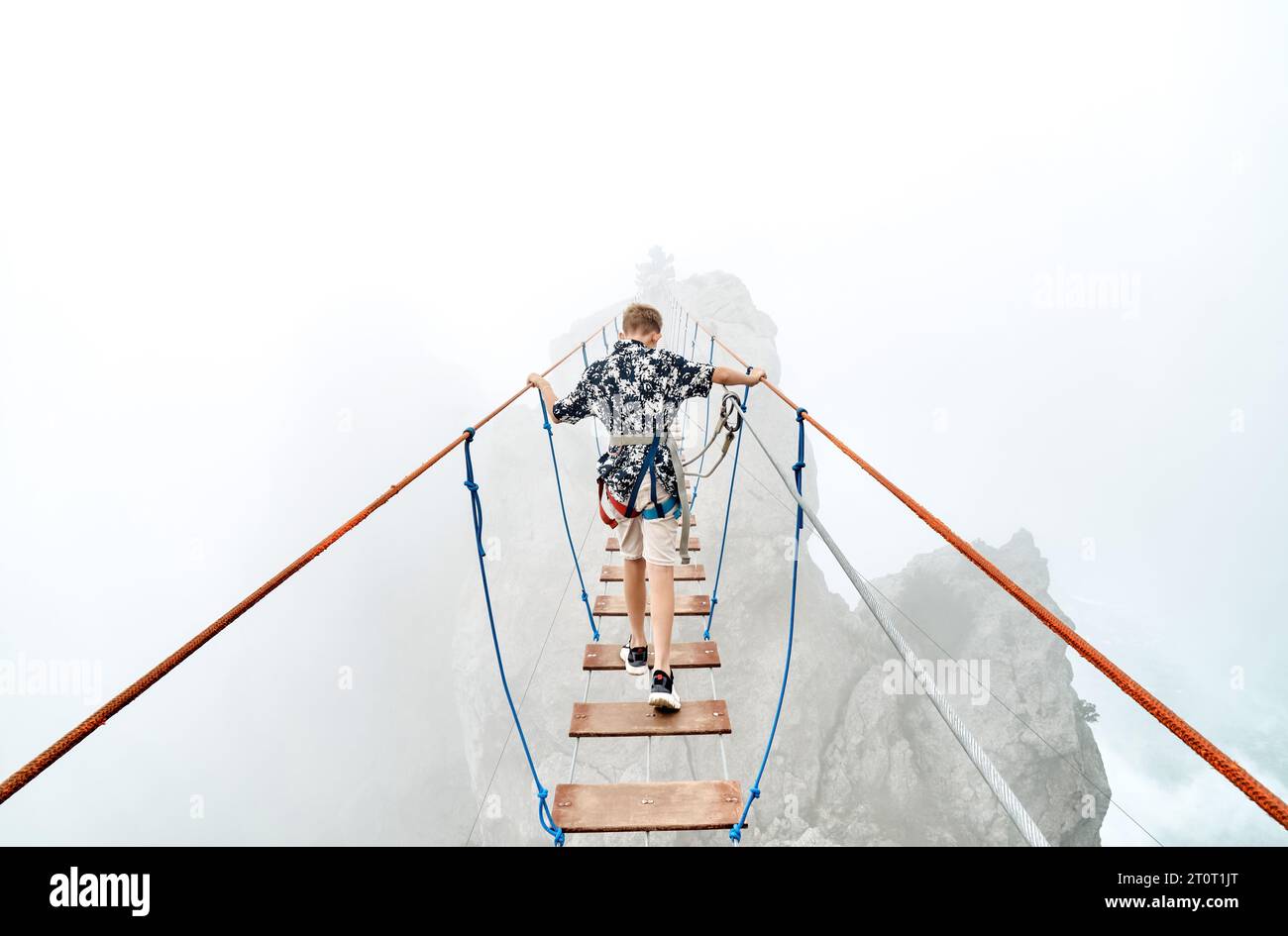 Courageous boy walks along rope bridge in mountain park on foggy day ...