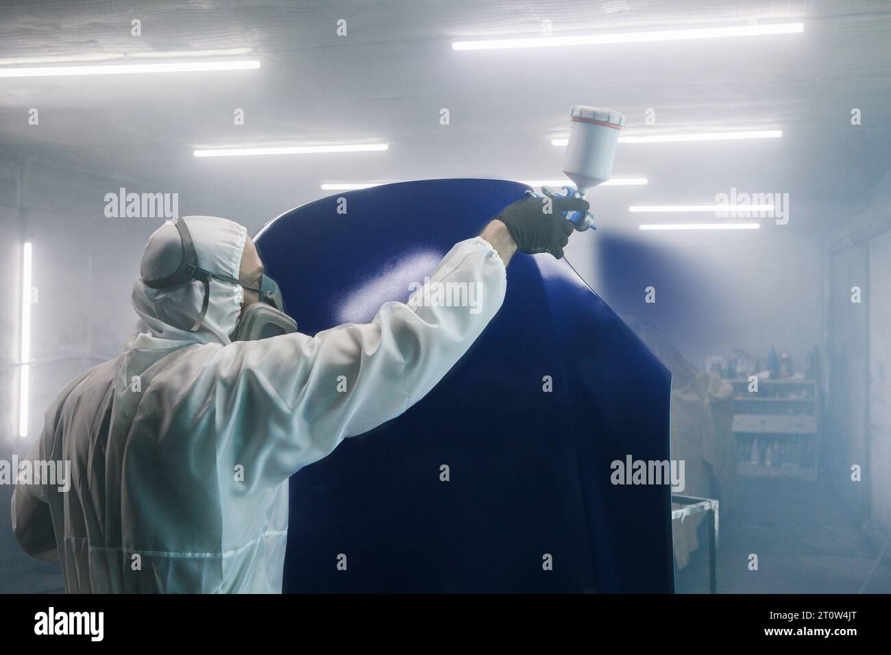 Worker paints car body part in workshop chamber using airbrush. Man ...