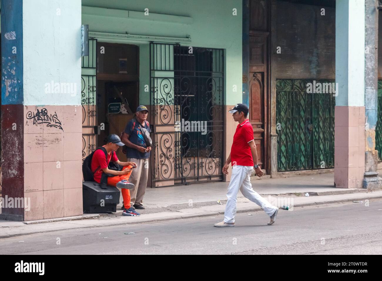 A Cuban man crosses a city street. Other people are below a porch in a ...