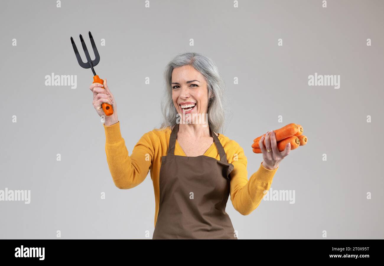 Cheerful senior lady farmer in apron rise up garden rake, show carrots ...