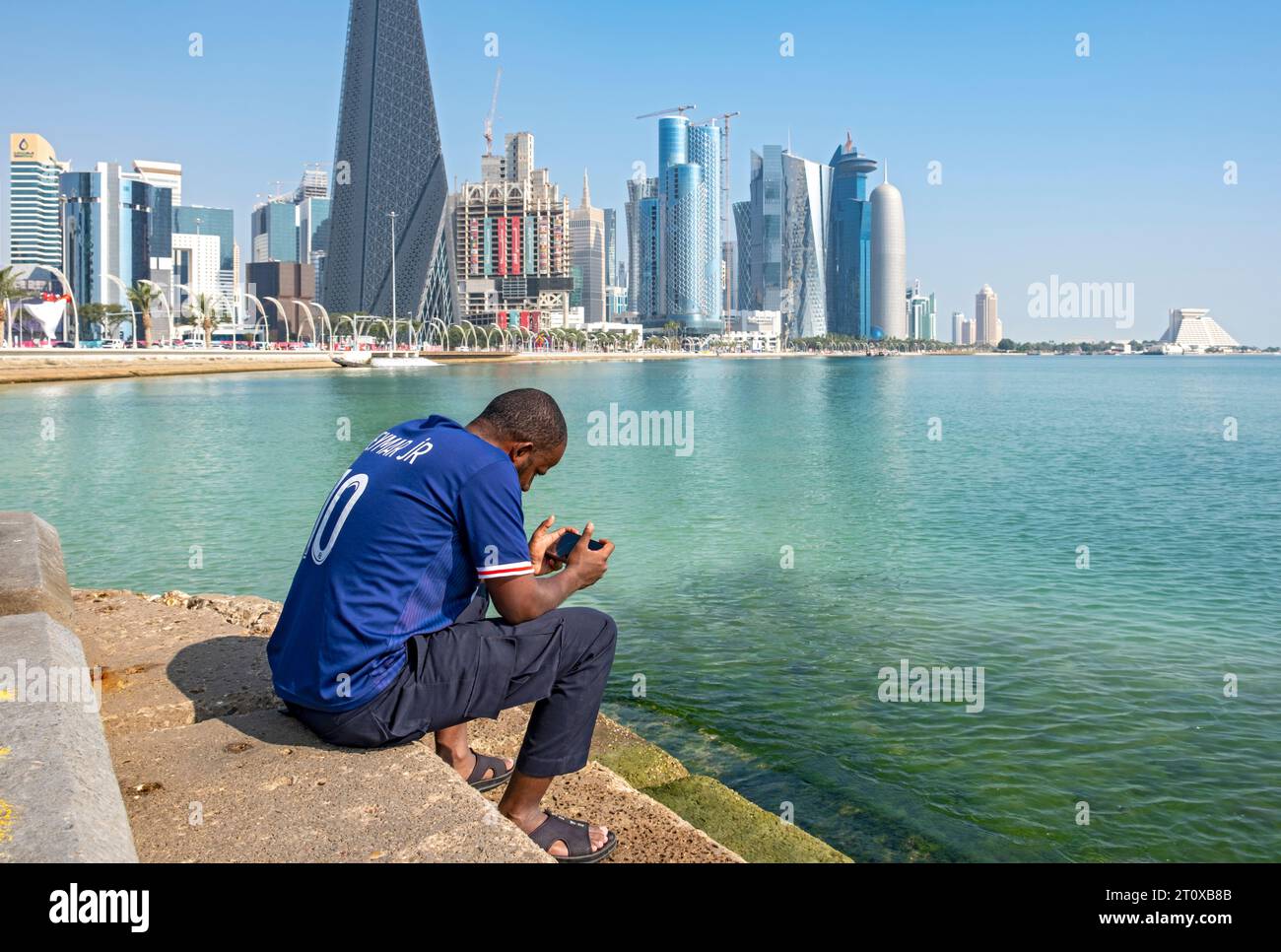 Man sits on steps of Corniche with skyscrapers in background, West Bay ...