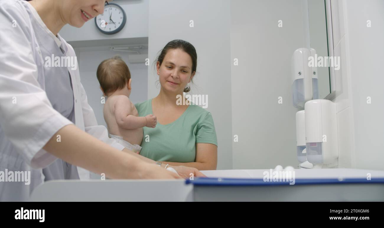 Mother holds baby on hands. Medic puts scale on changing table. Female ...