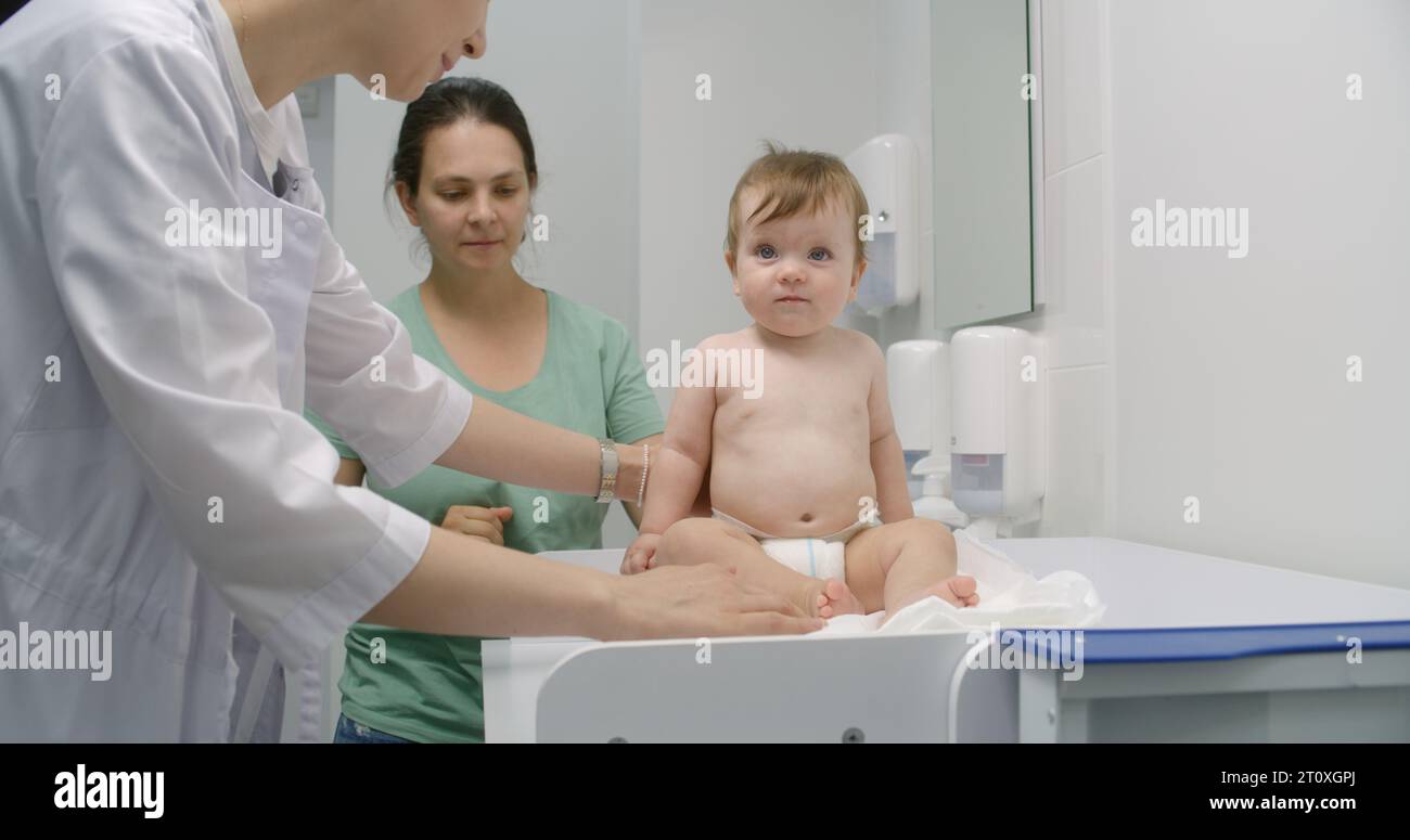 Mother holds baby on hands. Medic puts scale on changing table. Female ...