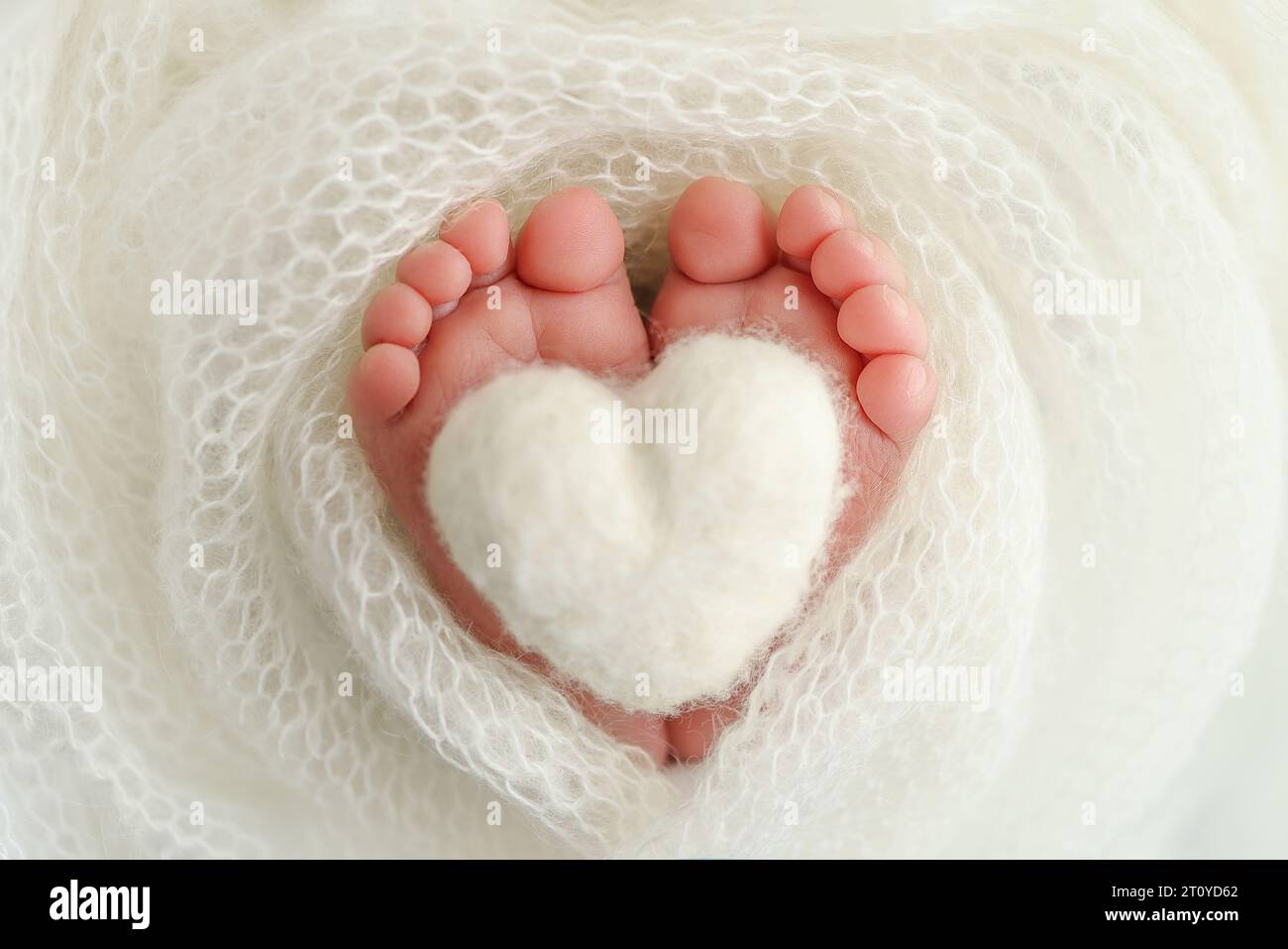 Knitted white heart in the legs of a baby. The tiny foot of a newborn ...
