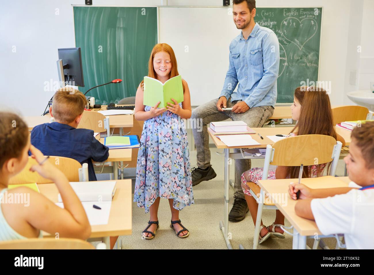 Smiling elementary student reading book with friends and teacher in ...
