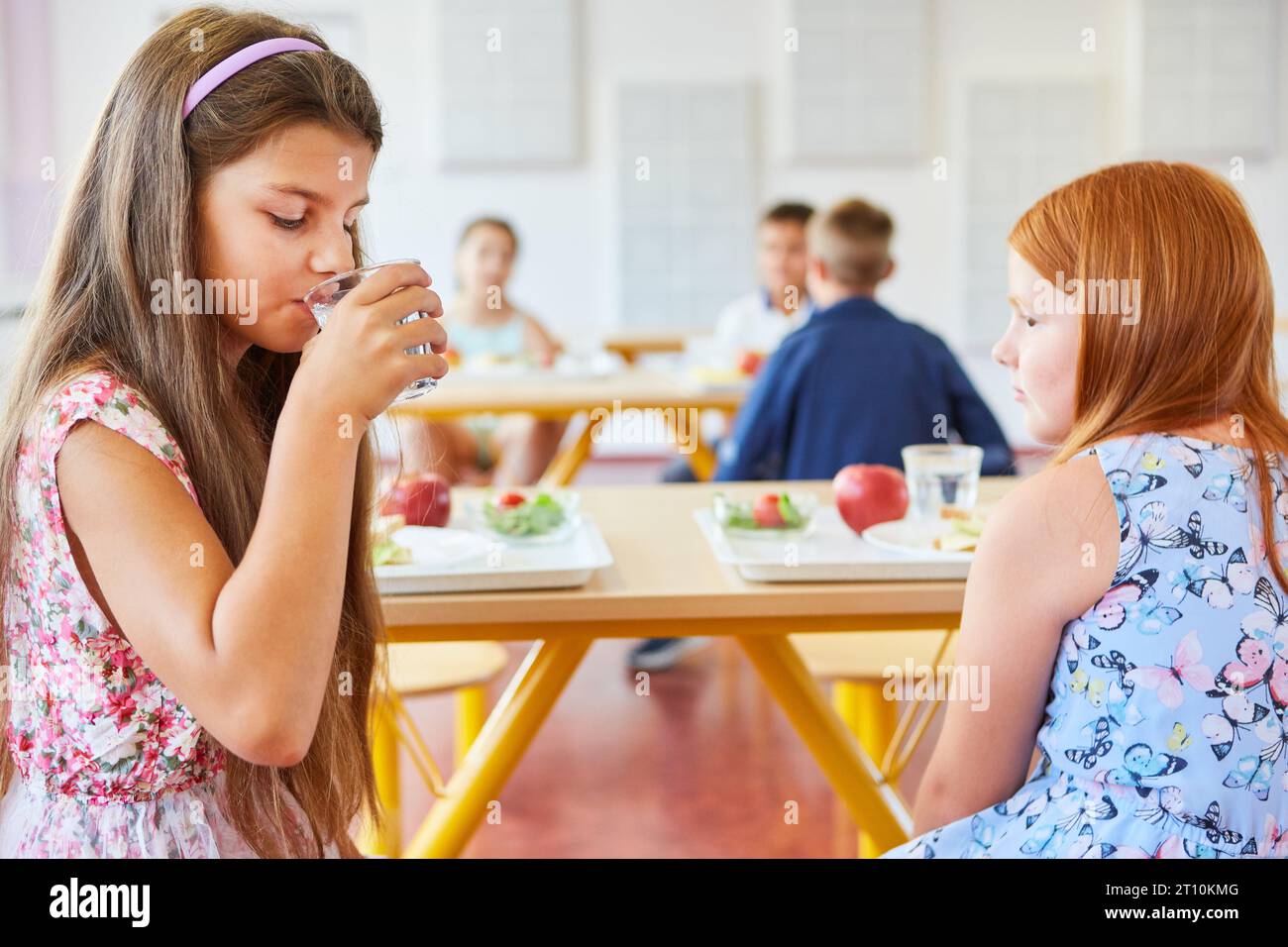 Thirsty girl drinking water from glass while sitting with female friend ...