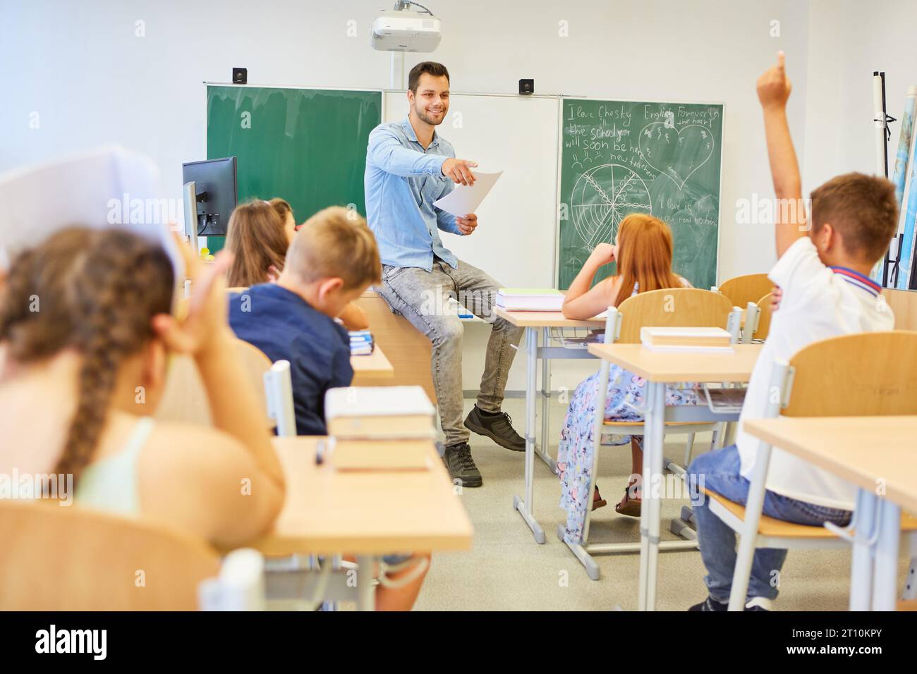Smiling male teacher pointing at student sitting with hand raised while ...