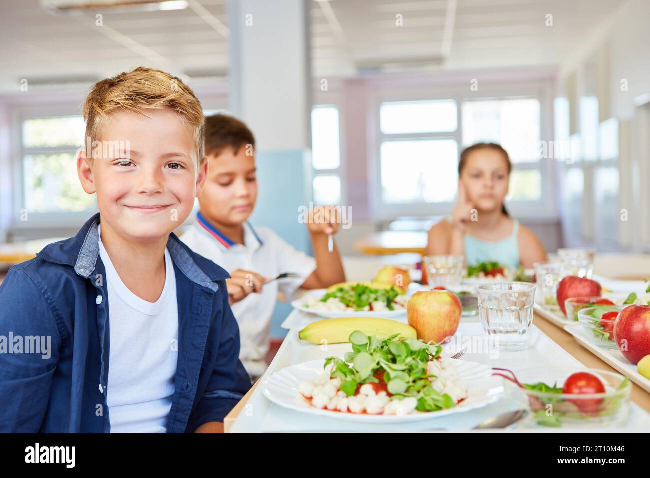 Portrait of smiling blond boy sitting with healthy food plate on table ...