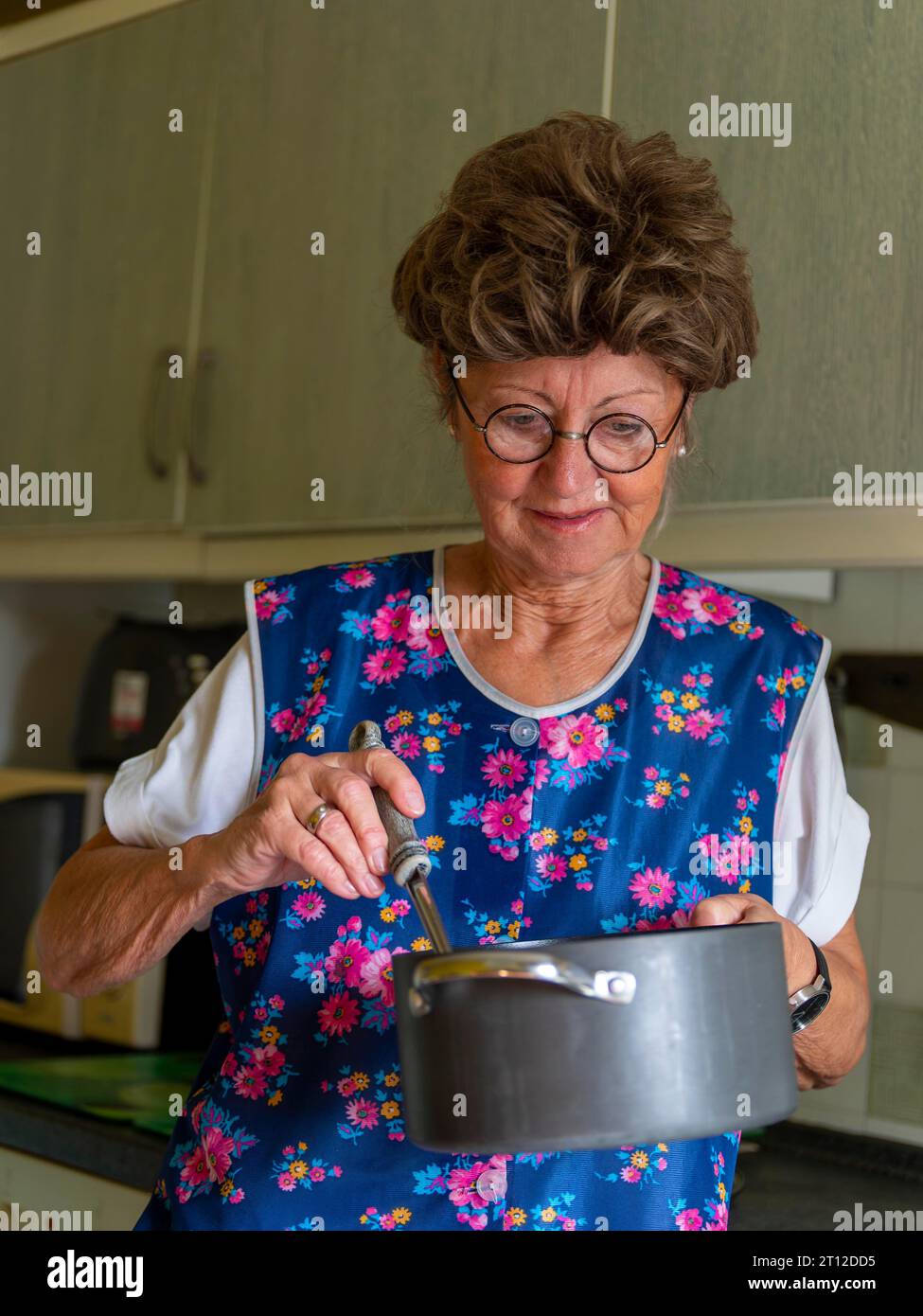 Granny with old smock apron, glasses and wig in the kitchen, holding ...