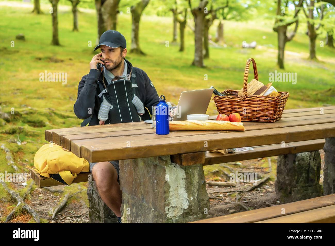 A young Caucasian father teleworking seated at a picnic table with the ...