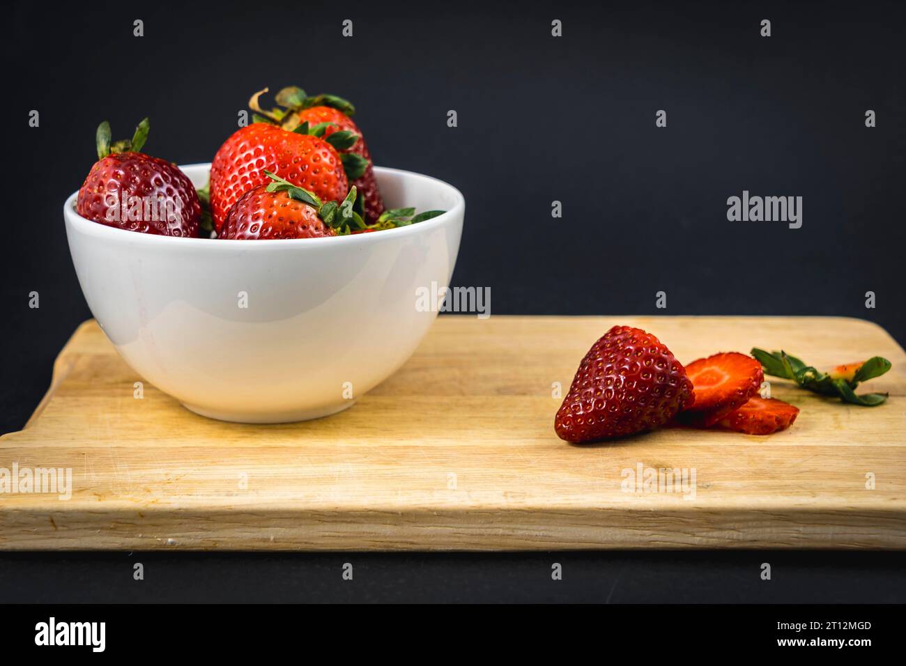 A strawberry cut on a wood and a white bowl with more strawberries ...