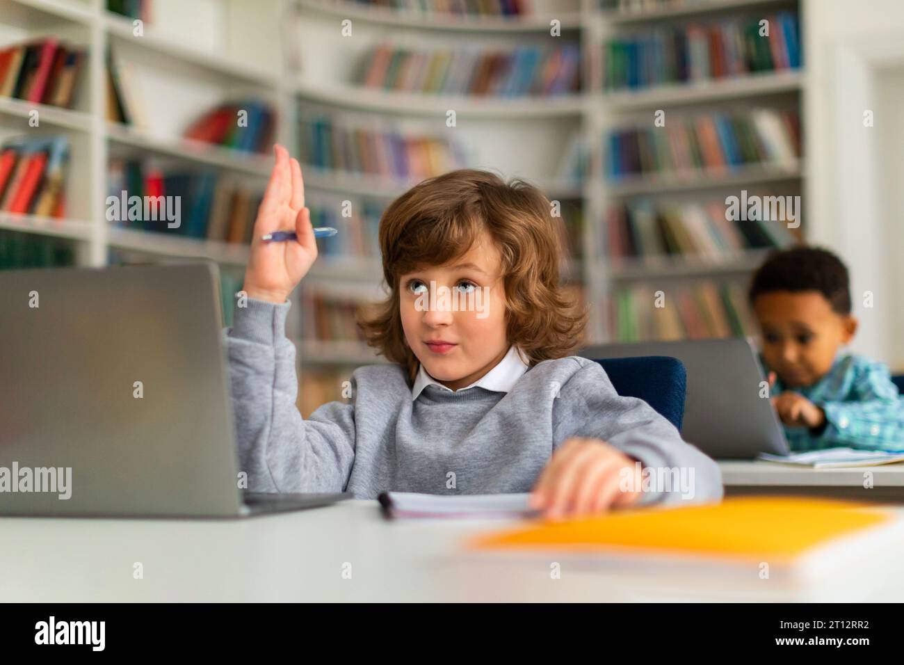 Schoolboy raising hand while using laptop in class Stock Photo - Alamy
