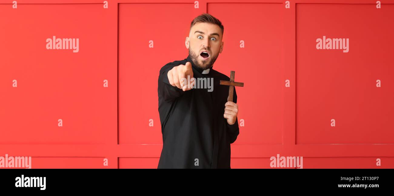 Shocked young priest with cross pointing at viewer against red ...