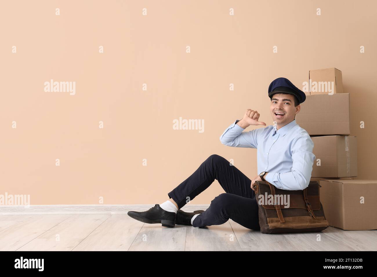Young postman with bag and parcels pointing at himself near beige wall ...