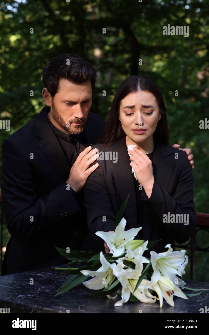 Sad couple mourning near granite tombstone with white lilies at ...