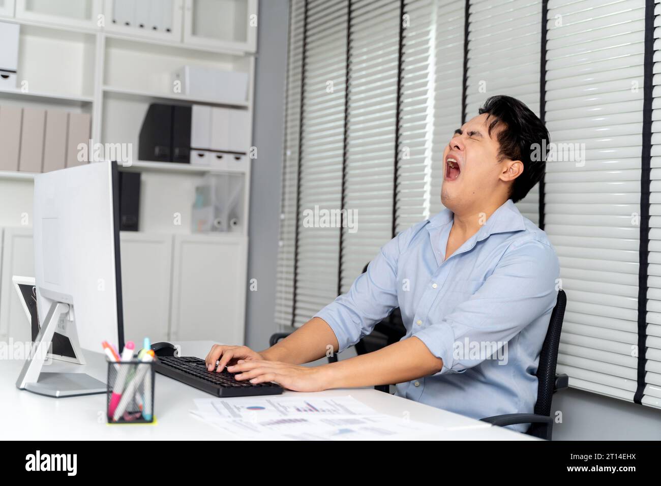 Young adult Asian businessman yawning on his desk with a computer pc in ...