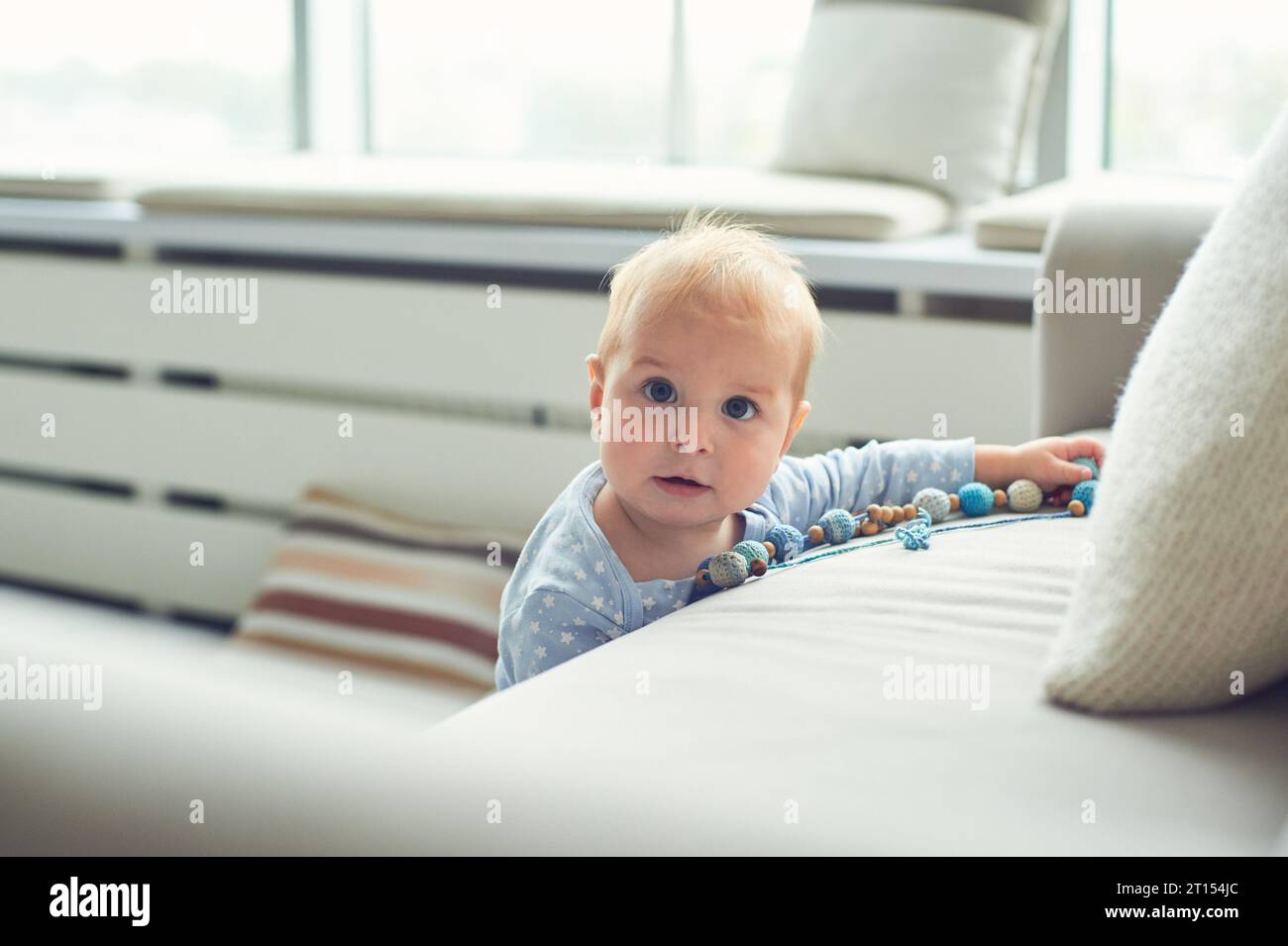 Little 8 months baby boy stands with support near sofa at home in white ...