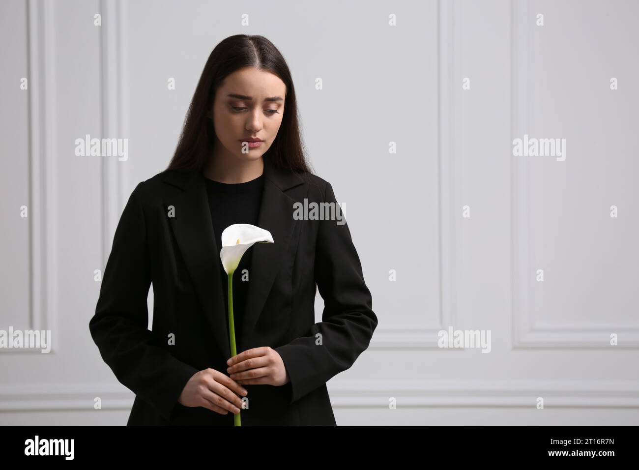 Sad woman with calla lily flower near white wall, space for text ...