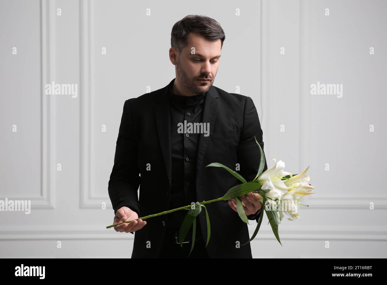 Sad man with lily flowers near white wall. Funeral ceremony Stock Photo ...