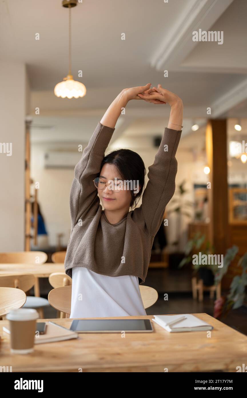 A relaxed and happy young Asian woman is stretching her arms, resting ...