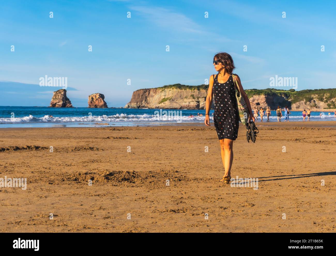 A young brunette in a black dress and sunglasses walking along the ...