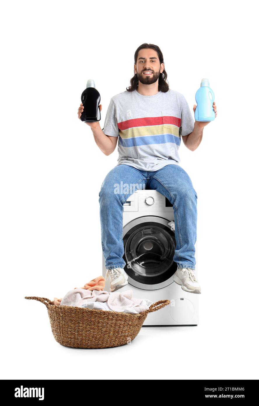 Young man with laundry detergents sitting on washing machine against ...