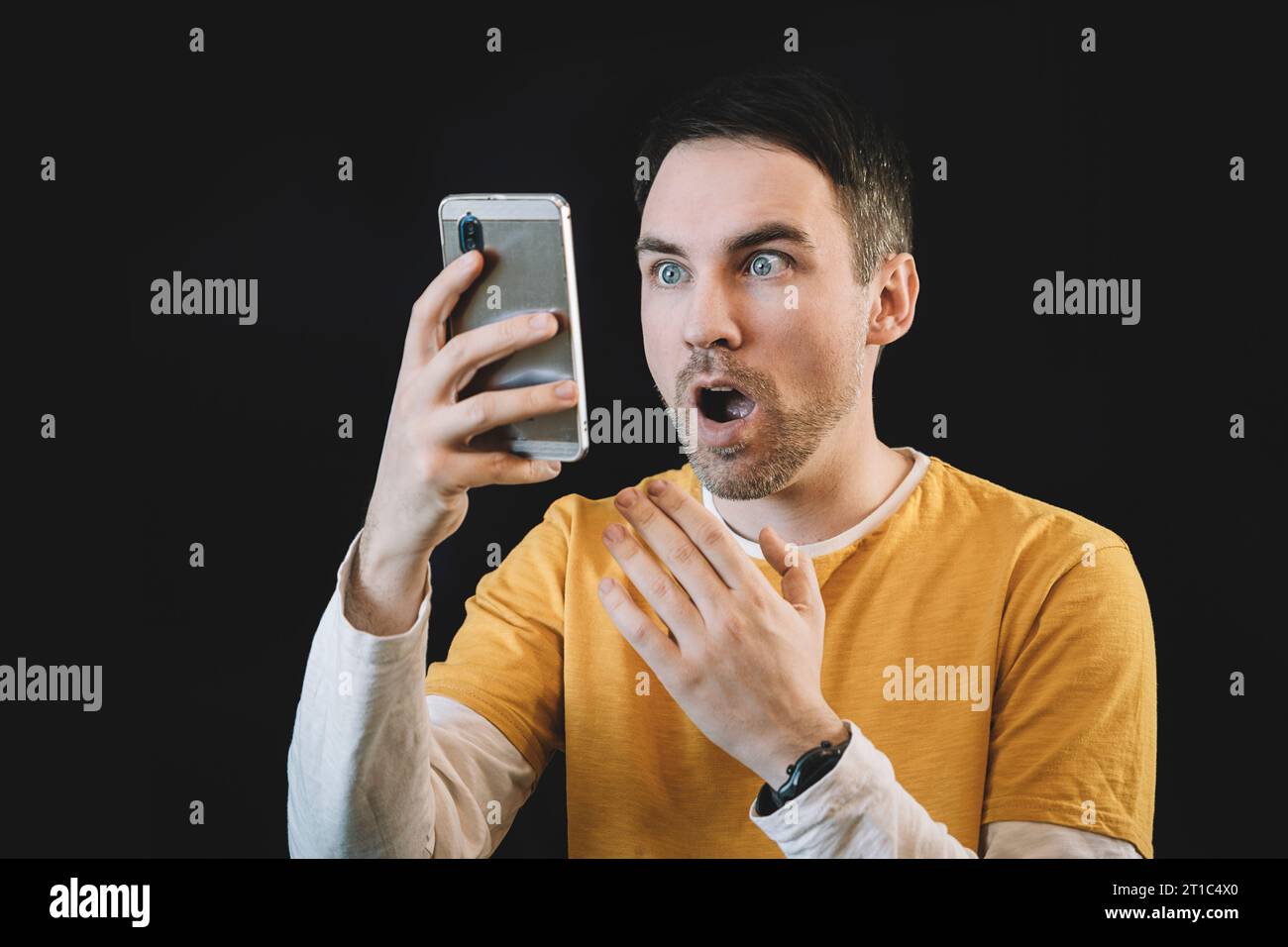 Shocked Young Caucasian man wearing pink black background open mouth hold phone reading advert ...