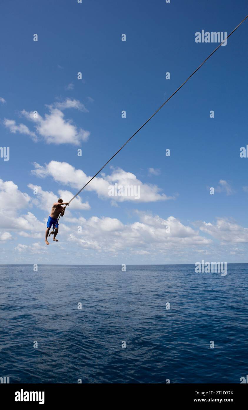 A man swings from a rope over the ocean against a blue sky with white ...