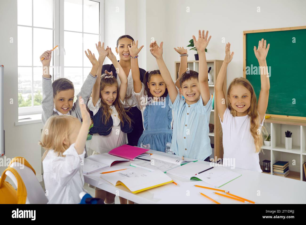 Happy children and young teacher are standing near desk rising hands up ...